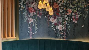 Um banco estofado verde com uma mesa redonda de madeira, um mural floral na parede e luminárias pendentes.