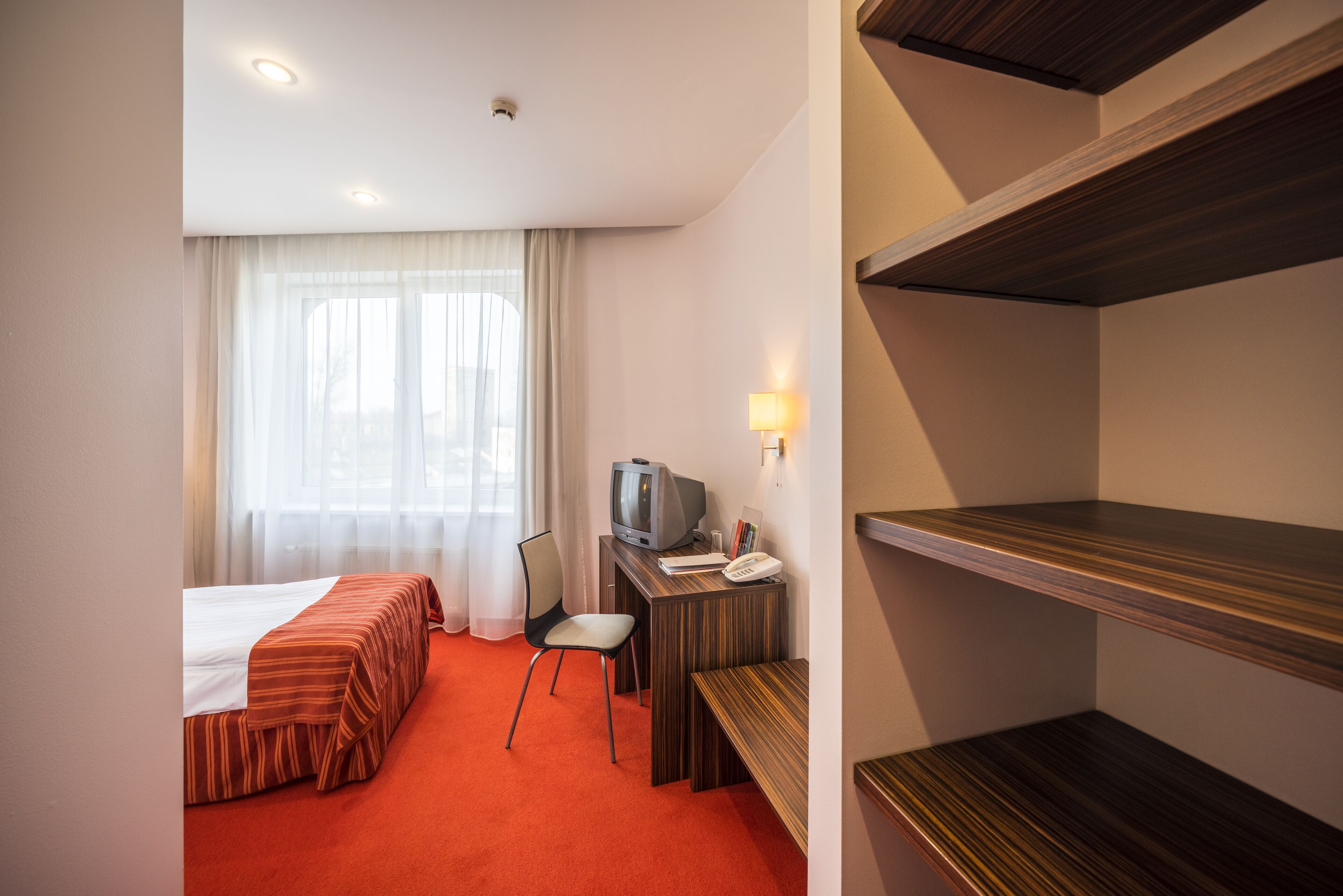 Quarto de hotel com cama, escrivaninha com televisão, cadeira e abajur.