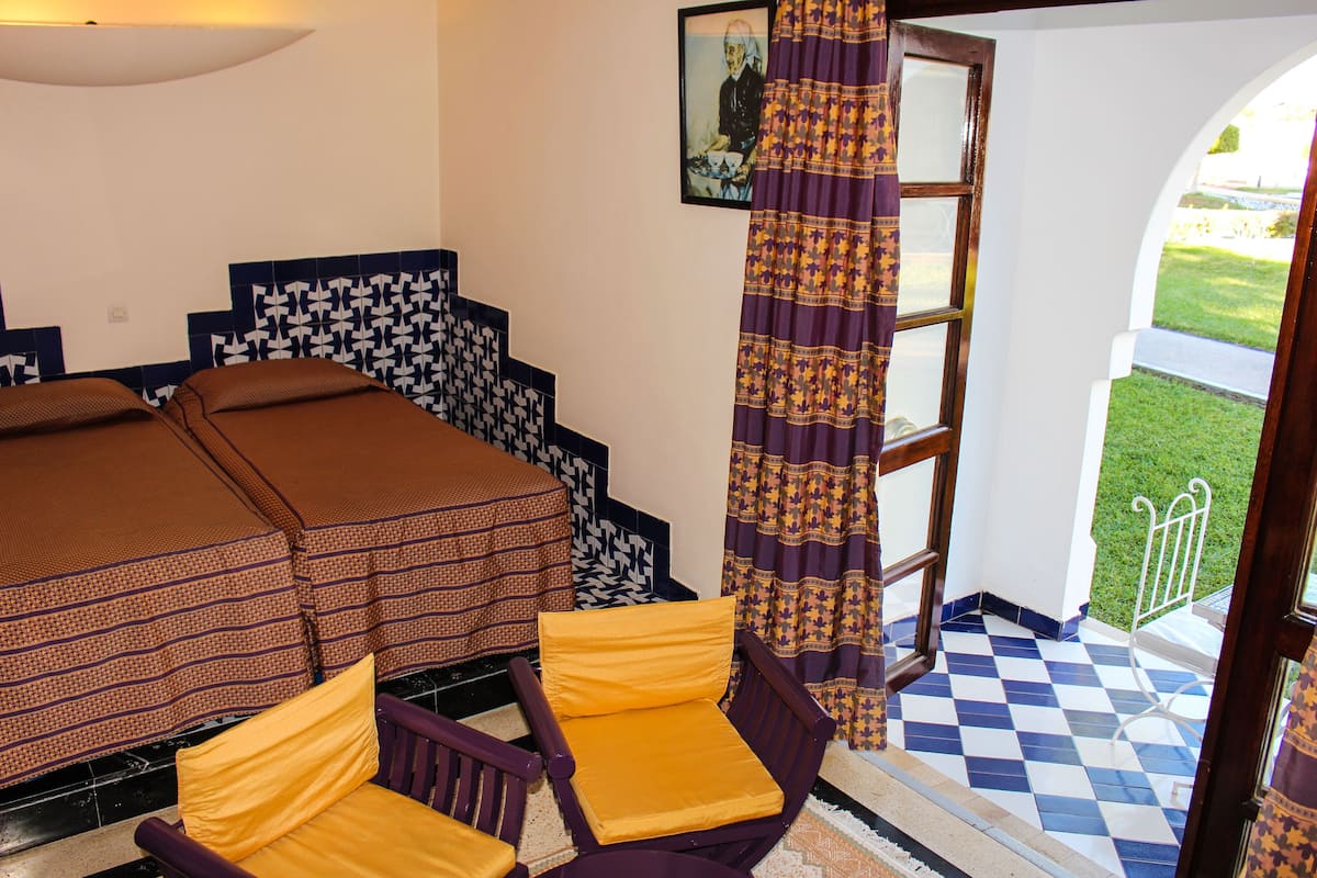 Quarto com uma cama, duas cadeiras, parede estampada, janela com cortinas e chão xadrez.