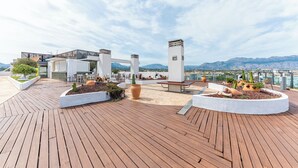 Terraço no telhado com piso de madeira, vasos brancos com plantas e vista para montanhas ao longe.