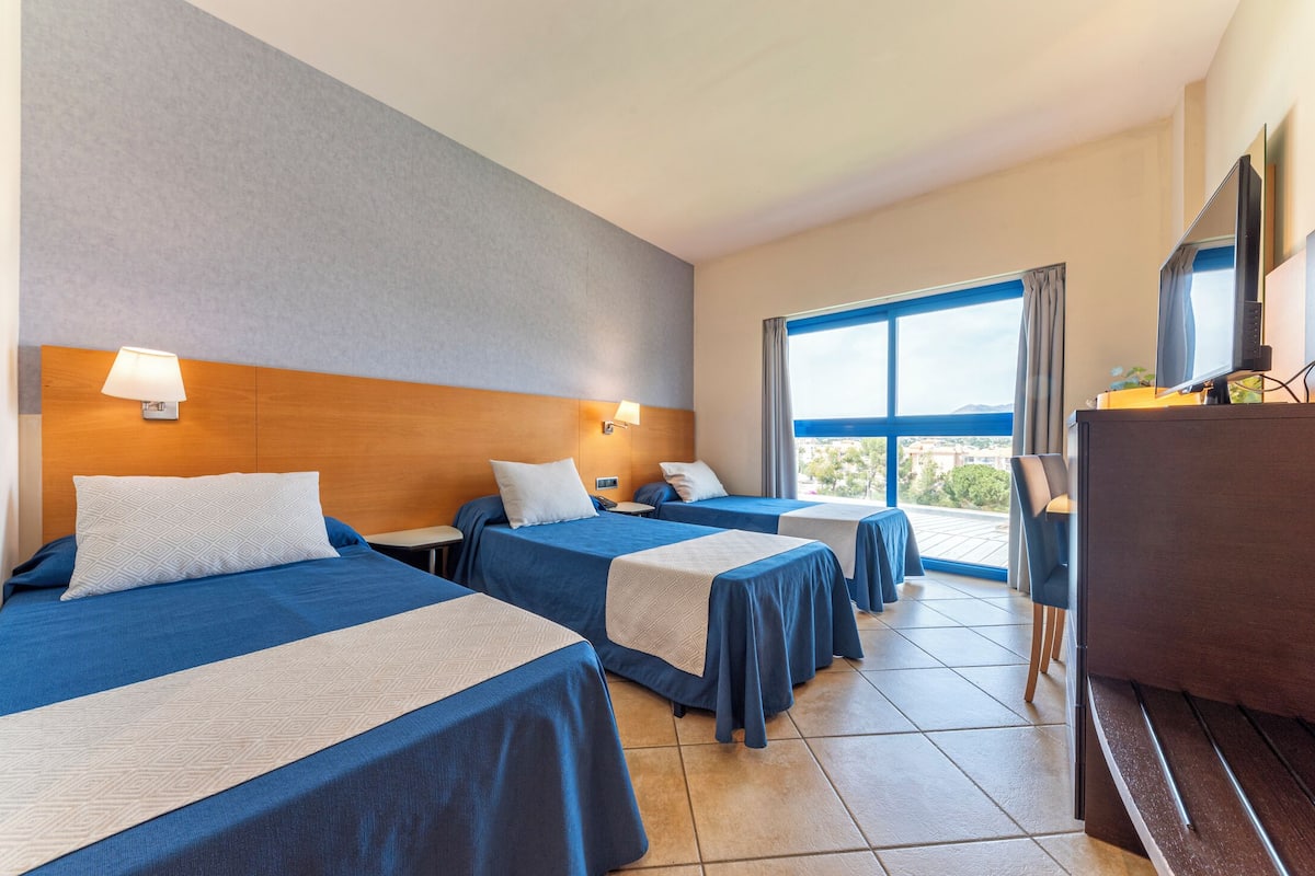 Quarto de hotel com duas camas, uma escrivaninha, televisão e uma grande janela com vista.