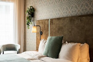Cama bem arrumada com almofadas, luminária fixada na parede e uma cadeira num quarto com papel de parede estampado.