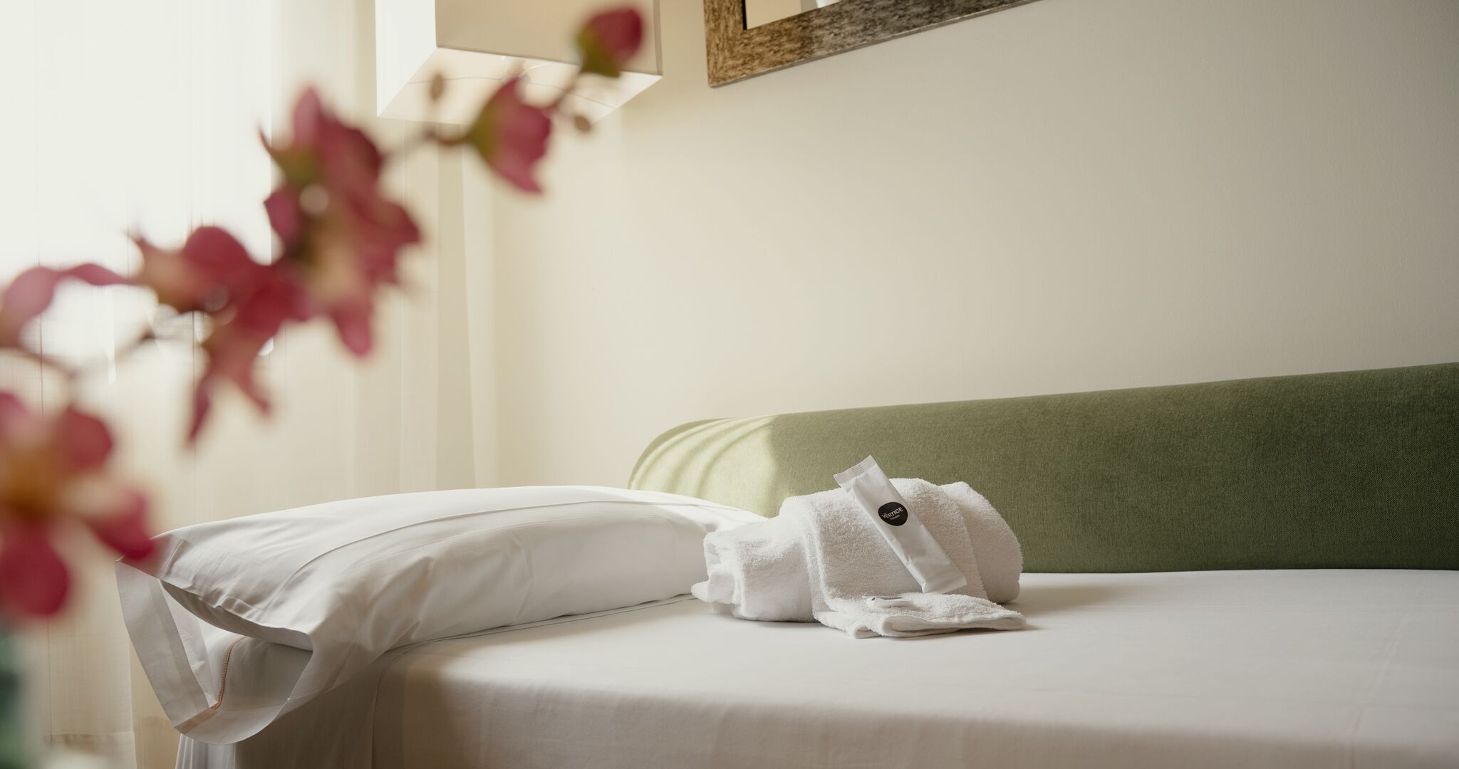 Cama bem arrumada com lençóis brancos e cabeceira verde, um travesseiro branco e uma toalha branca dobrada na cama.