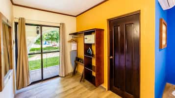 Quarto de hotel com piso de madeira, uma porta, uma pequena prateleira com objetos e vista para um jardim através de uma porta de vidro.