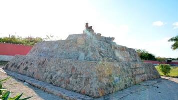 Uma estrutura piramidal de pedra com uma estátua no topo, cercada por um espaço pavimentado e uma parede vermelha ao fundo.