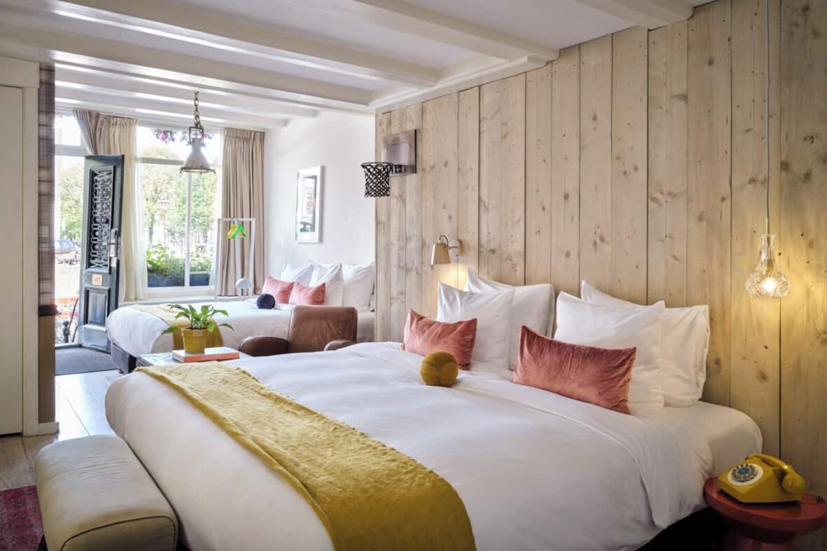 Quarto de hotel com uma cama grande, parede de madeira, uma mesa pequena com telefone e uma janela com cortinas.
