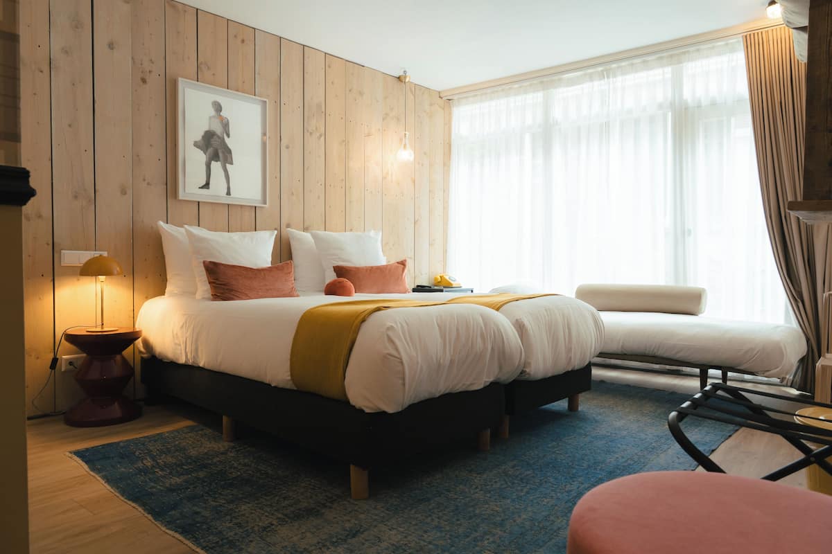Quarto de hotel com parede de madeira, cama com roupa de cama branca e rosa, tapete azul e janela com cortinas leves.