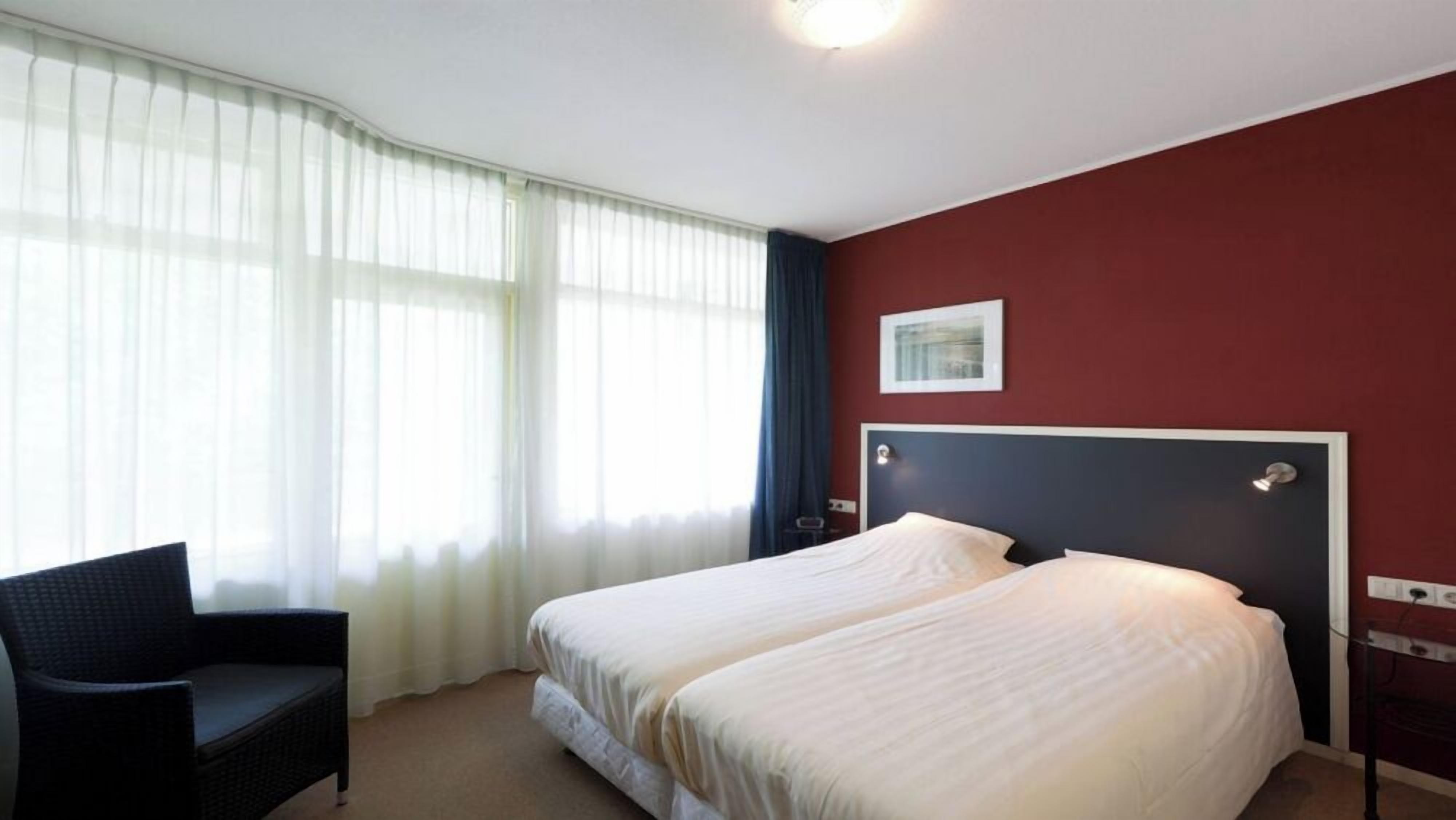 Quarto de hotel com cama, cadeira, janela com cortinas e quadro na parede.