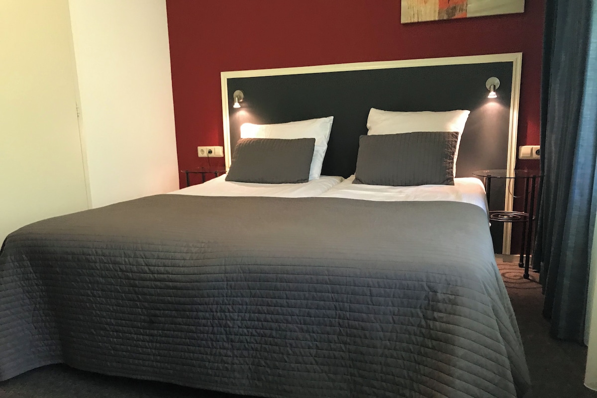 Cama bem arrumada com um edredom escuro, cabeceira com duas luzes fixadas na parede, e uma mesinha de cabeceira com abajur ao lado da cama.