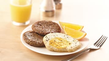 Um prato com duas hambúrgueres, um ovo frito e uma fatia de laranja em um prato branco, acompanhado por um garfo e um copo de suco de laranja.