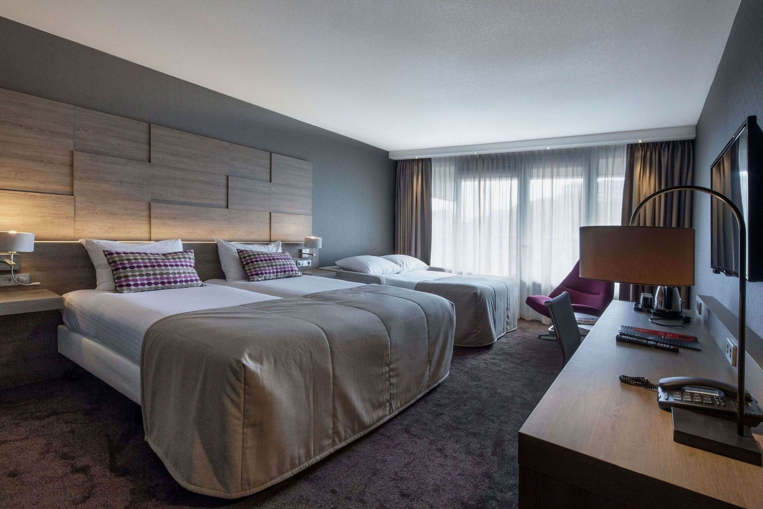 Quarto de hotel com duas camas, uma mesa de trabalho com luminária, uma cadeira e uma televisão.