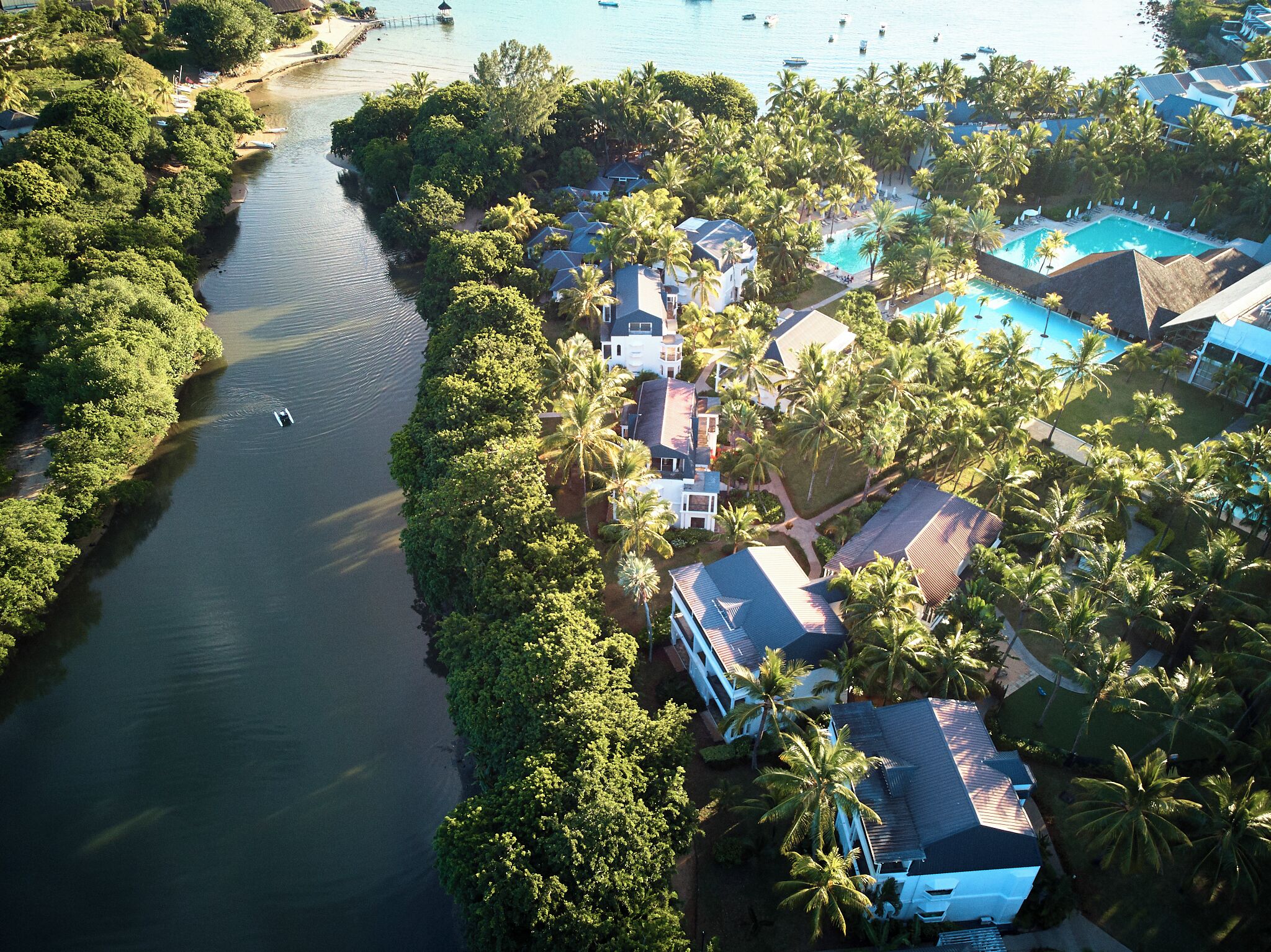 Vista aérea de um resort à beira-mar, com um rio, palmeiras e área de piscina.