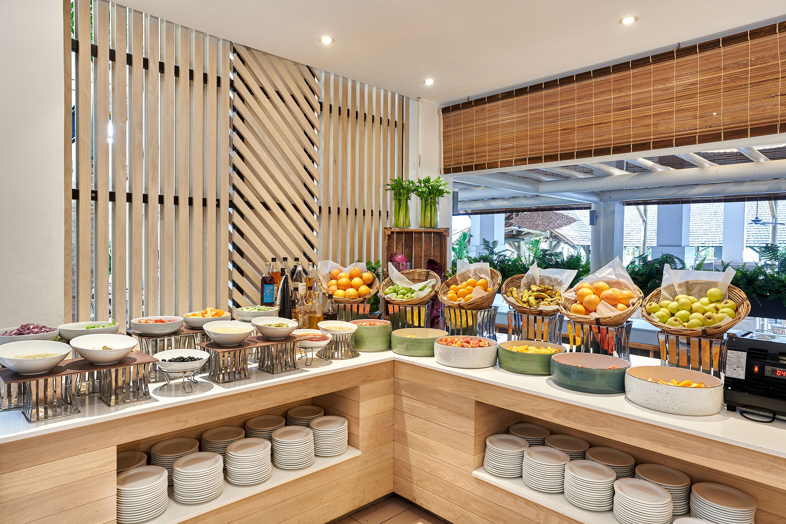 Disposição de buffet com diversos alimentos e recipientes, painéis de madeira e vista para o verde lá fora.