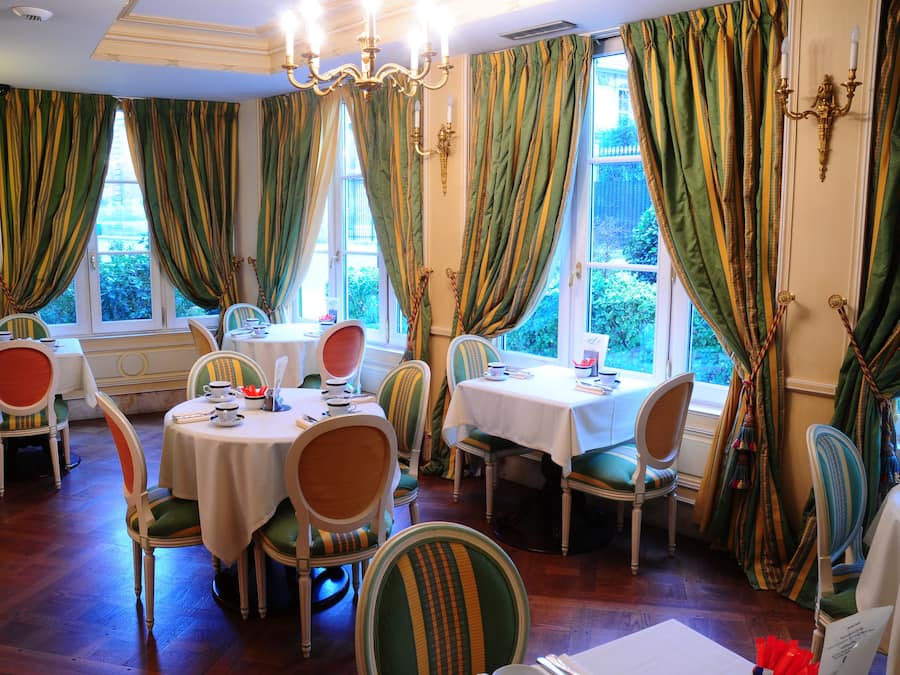 Uma área de refeições com mesas e cadeiras redondas, grandes janelas com cortinas e um lustre.