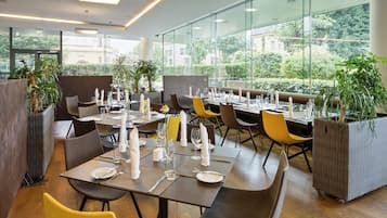 Uma área de jantar moderna com mesas para quatro pessoas, grandes janelas com vista para vegetação e edifícios, e plantas em vasos que trazem um toque de natureza.
