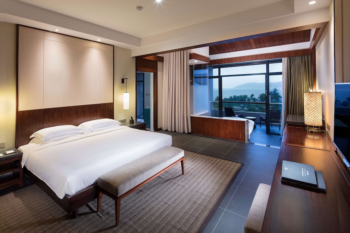 Quarto de hotel moderno com uma cama grande, uma escrivaninha e vista para o horizonte urbano através de grandes janelas.