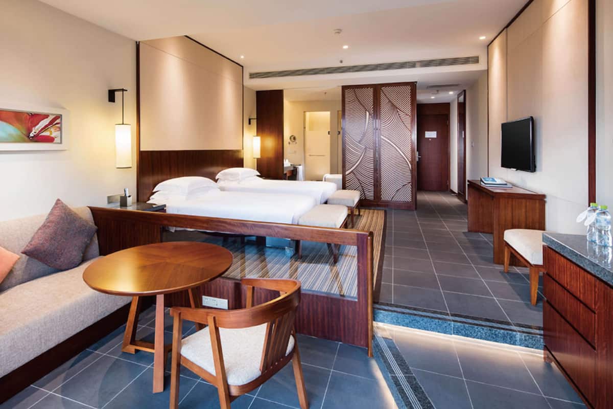 Quarto de hotel com sofá, duas camas, mesa de jantar, televisão e banheiro visível ao fundo.