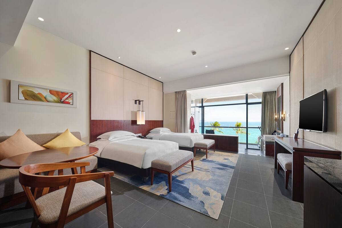 Quarto de hotel com duas camas, uma mesa, uma televisão e uma grande janela com vista para o oceano.