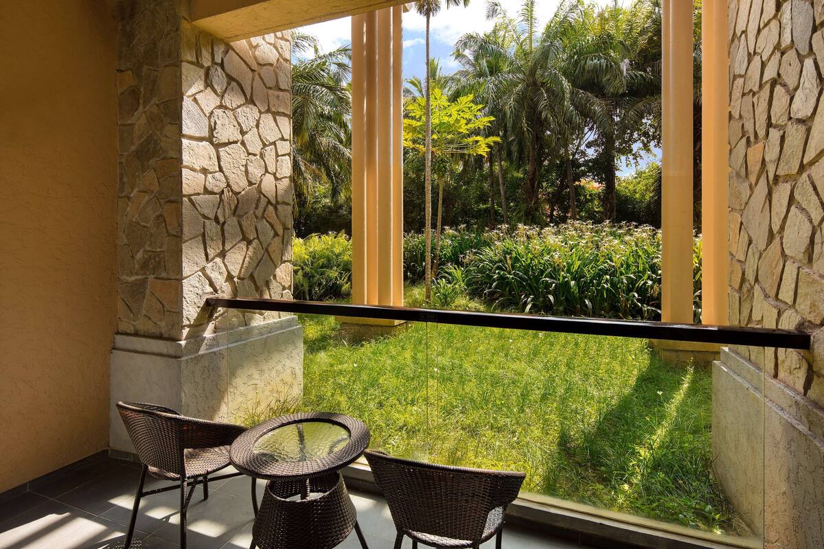 Varanda com cadeiras de vime e mesa de vidro, com vista para um jardim com palmeiras e um muro de pedra.