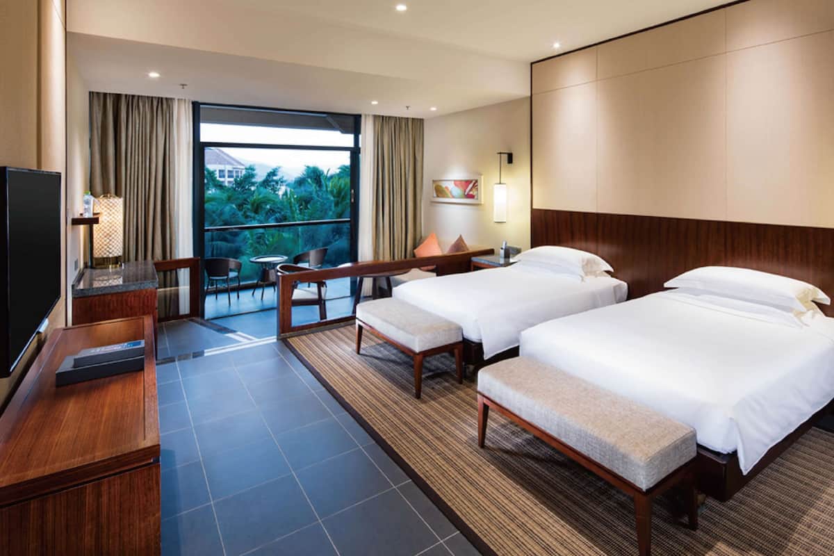 Quarto de hotel com duas camas, uma escrivaninha, uma televisão e vista para o verde lá fora.