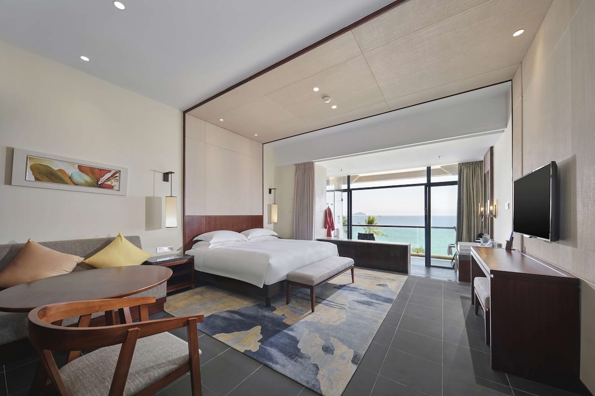 Quarto de hotel moderno com uma cama grande, uma escrivaninha, televisão e varanda com vista para o mar.
