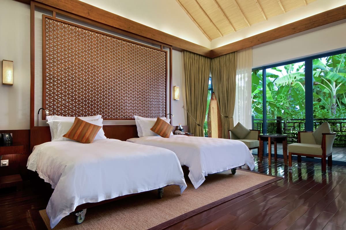 Quarto de hotel com duas camas, teto de madeira, janela ampla com vista para área verde e um painel decorativo tradicional.