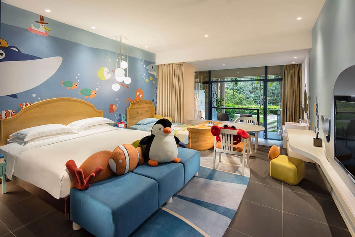 Quarto de hotel com cama, sofá azul, cabeceira de madeira, mesa de jantar com cadeiras, televisão e uma janela grande com vista para vegetação.