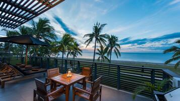 Um deck de madeira com mesa e cadeiras, com vista para uma praia com palmeiras e o oceano.