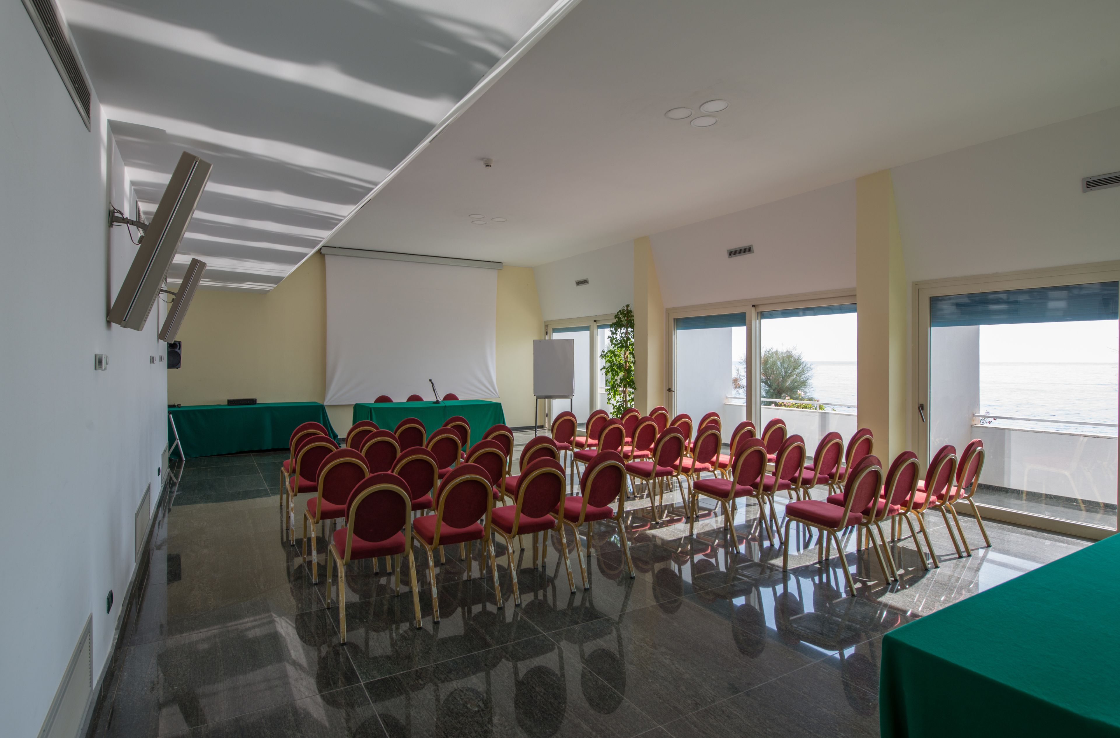 Uma sala de conferências com cadeiras vermelhas, uma tela de projetor e uma mesa com toalha verde.