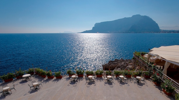 Um terraço à beira-mar com mesas ao ar livre e vista para o mar e uma montanha.