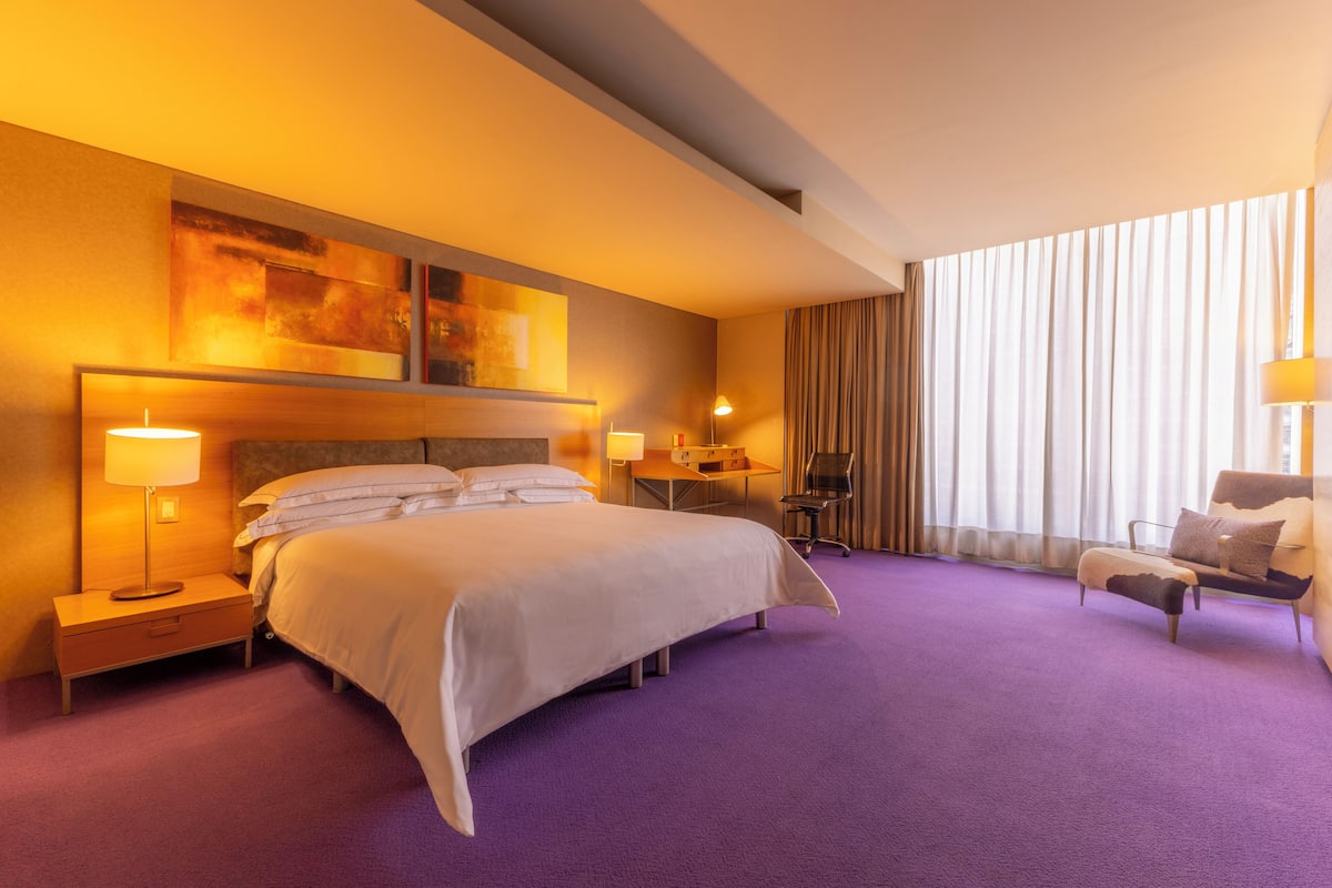 Quarto de hotel com uma cama grande, uma escrivaninha, uma cadeira e uma janela com cortinas.