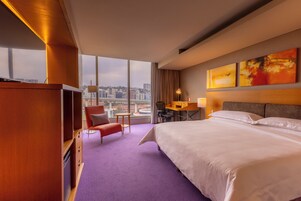 Quarto de hotel com cama grande, escrivaninha, cadeira e vista para a cidade.