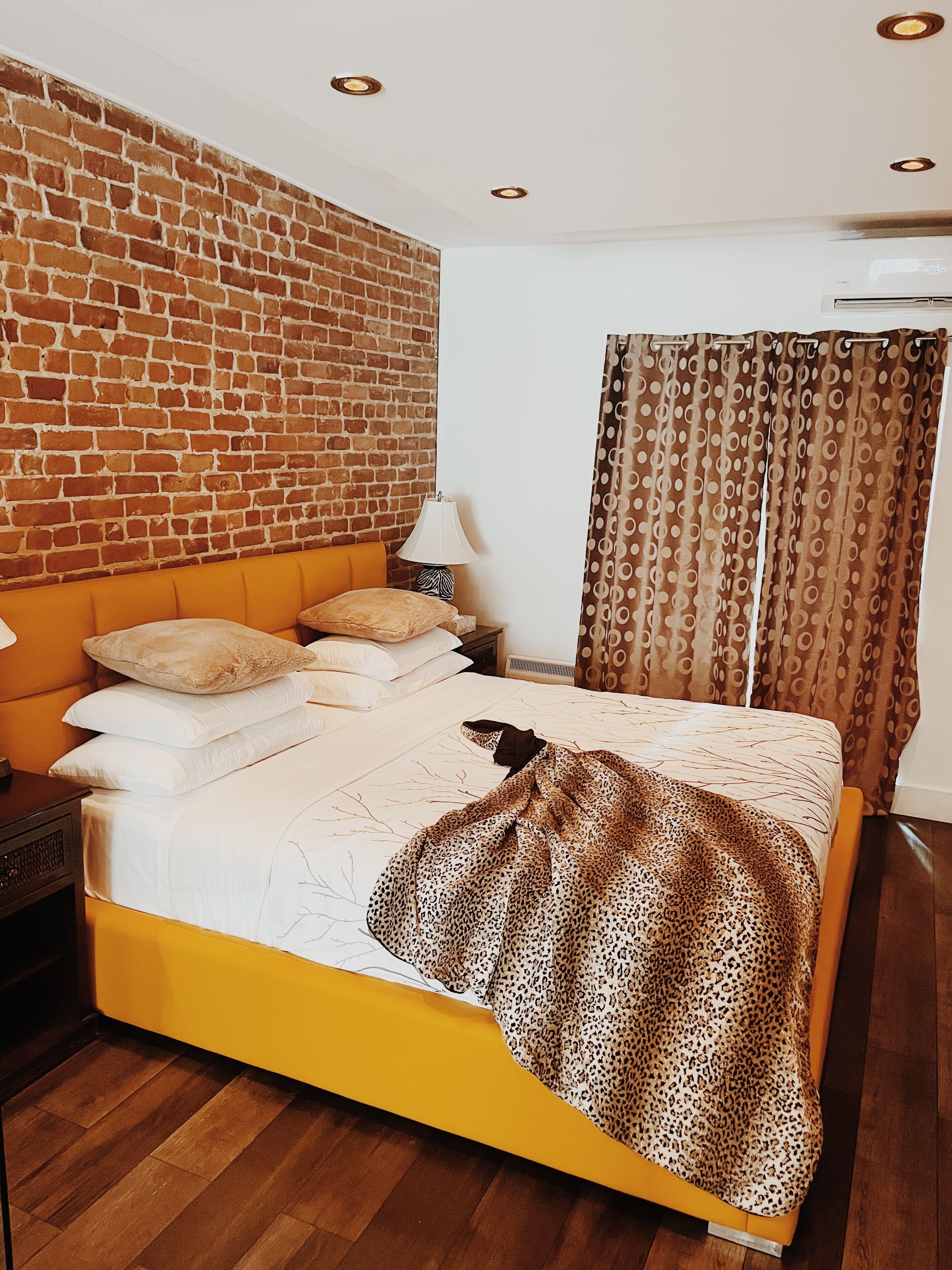 Quarto com parede de tijolos, cama com roupa de cama branca e manta de onça, cabeceira laranja e mesa de cabeceira com abajur.