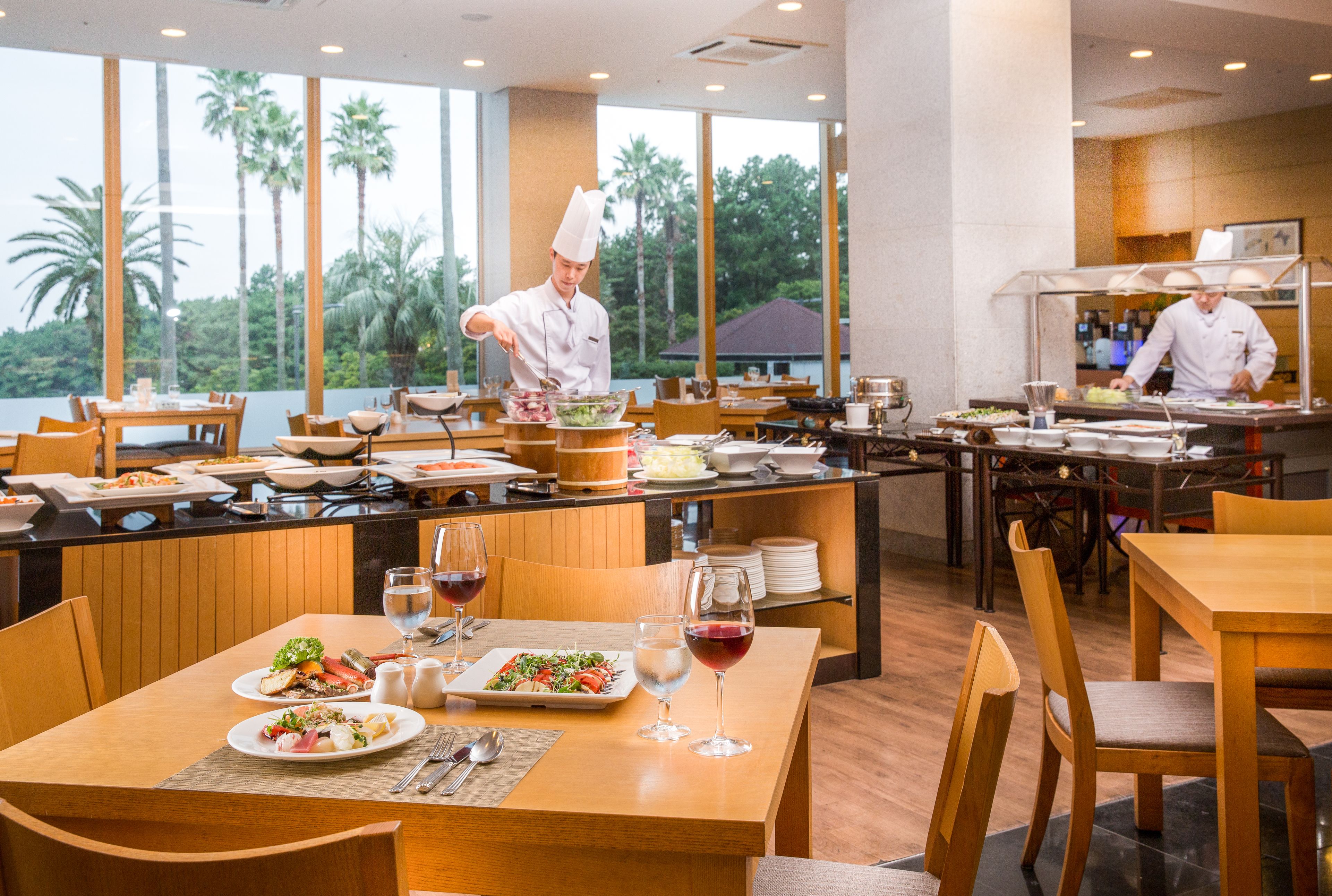 Restaurante com vista para palmeiras, mesas de madeira e um chef preparando refeições.