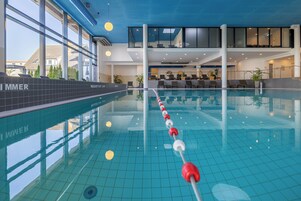 Piscina interior com água cristalina, bloco de partida com boias demarcando as pistas e área de recepção com assentos e balcão.