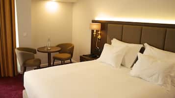 Quarto de hotel com uma cama grande, duas poltronas, uma mesinha redonda e uma mesa de cabeceira com abajur.