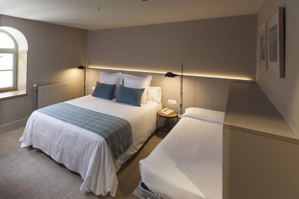 Quarto de hotel com duas camas, uma janela, um radiador e uma mesa de cabeceira com abajur.
