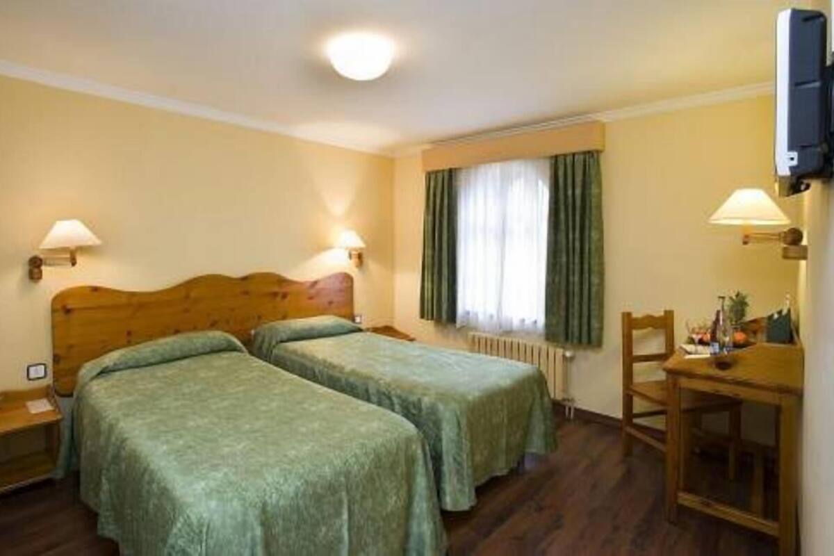 Quarto de hotel com duas camas de solteiro, cabeceira de madeira, uma mesa pequena com uma cadeira e uma janela com cortinas.