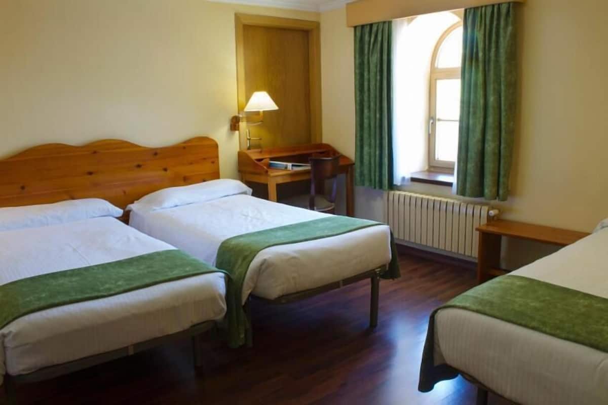Quarto de hotel com duas camas, uma mesa, uma luminária e uma janela com cortinas.