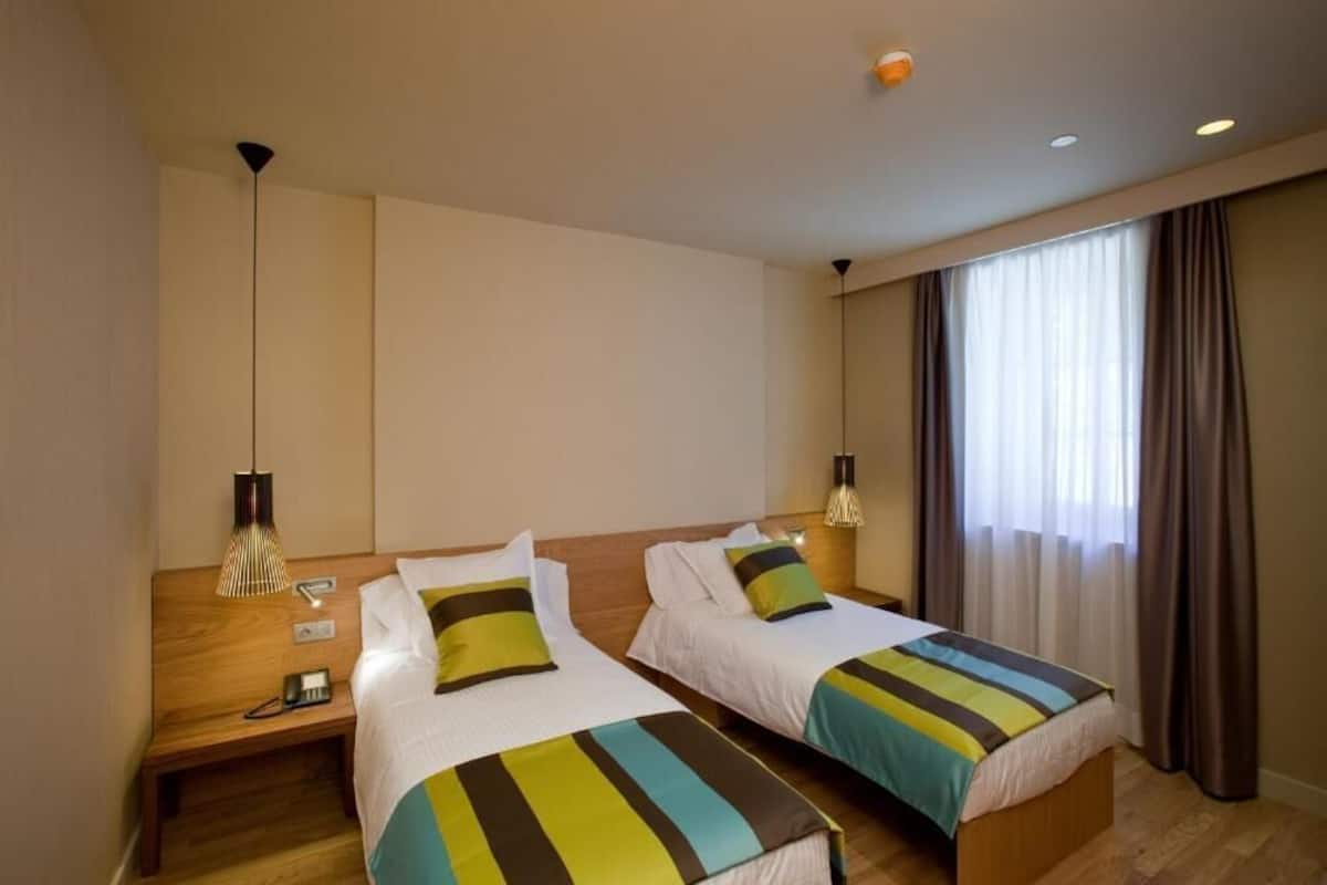 Quarto de hotel com duas camas de solteiro, cabeceiras de madeira, mesas de cabeceira com abajures e uma janela grande com cortinas.