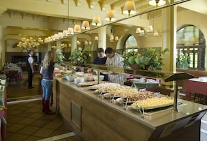 Uma área de buffet com várias estações de comida, uma pessoa servindo e um garçom ao fundo.