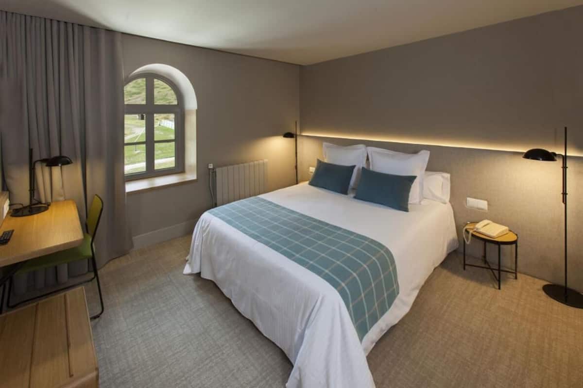 Quarto de hotel com cama, escrivaninha, cadeira, luminária, mesa de cabeceira e janela.