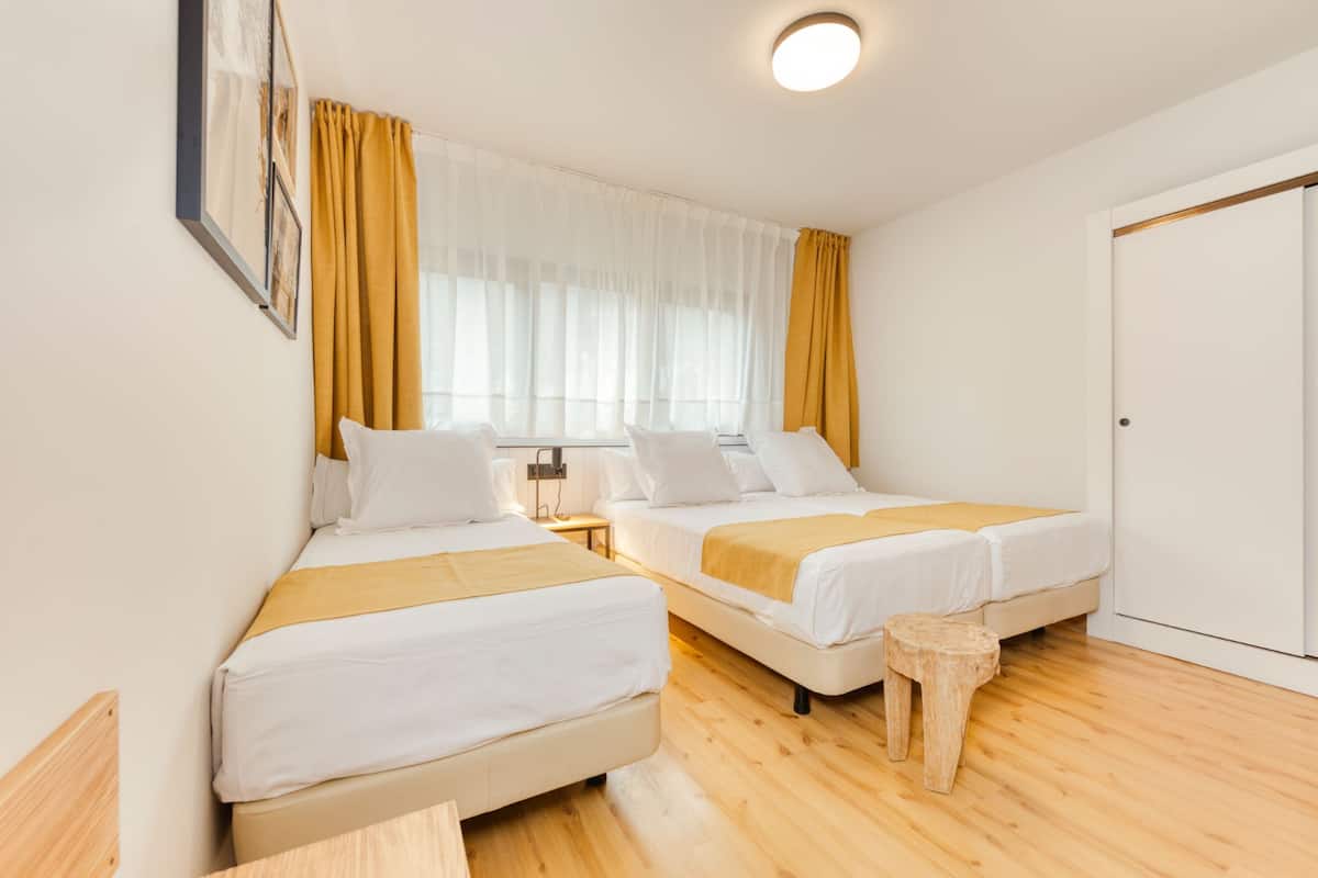 Quarto de hotel com duas camas, piso de madeira, um banquinho e uma porta.