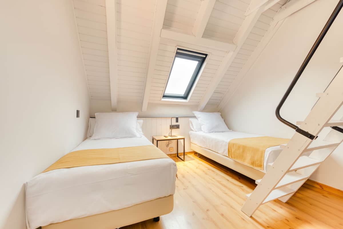 Quarto pequeno e bem iluminado, com duas camas, um claraboia e piso de madeira.