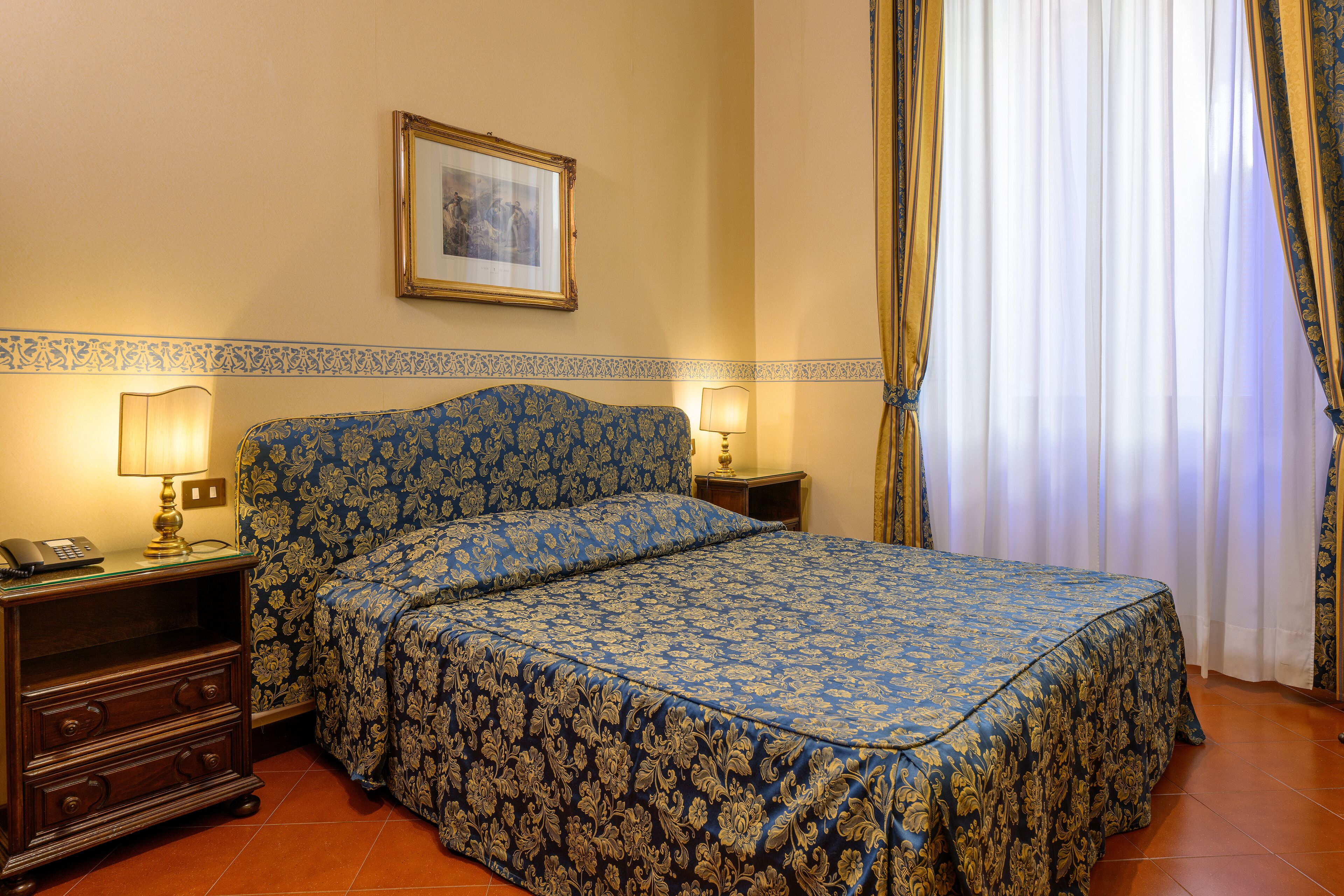 Quarto com uma cama grande, duas mesas de cabeceira, um criado-mudo, um quadro emoldurado na parede e uma janela com cortinas.