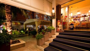 Um saguão de hotel com escada que leva à área do bar, plantas dentro de casa e uma área de estar com mesas e cadeiras.