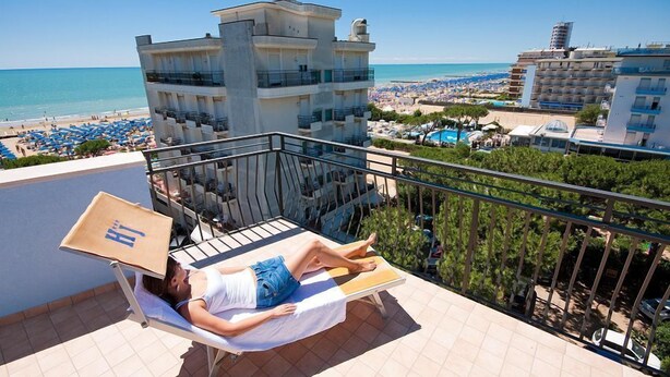 Varanda com uma pessoa tomando sol, com vista para uma praia com guarda-sóis e piscina.
