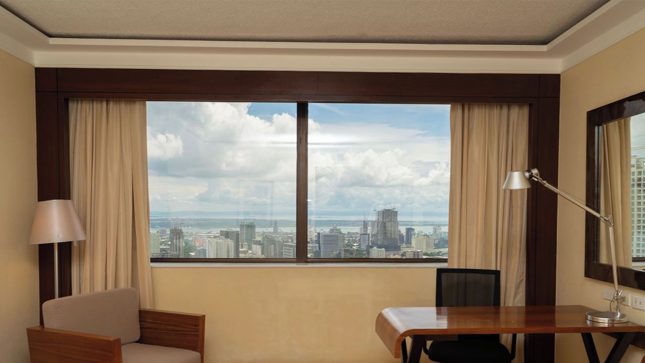 Quarto de hotel com vista para a cidade, uma escrivaninha, uma cadeira e um abajur.