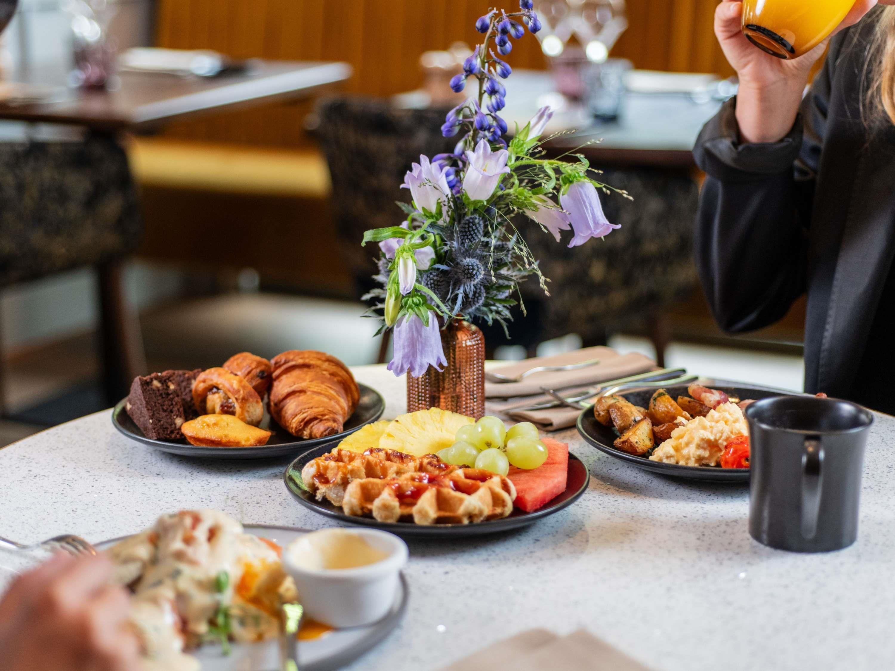 Um ambiente de café da manhã com croissants, waffles e frutas em pratos, um vaso de flores e uma xícara de café.