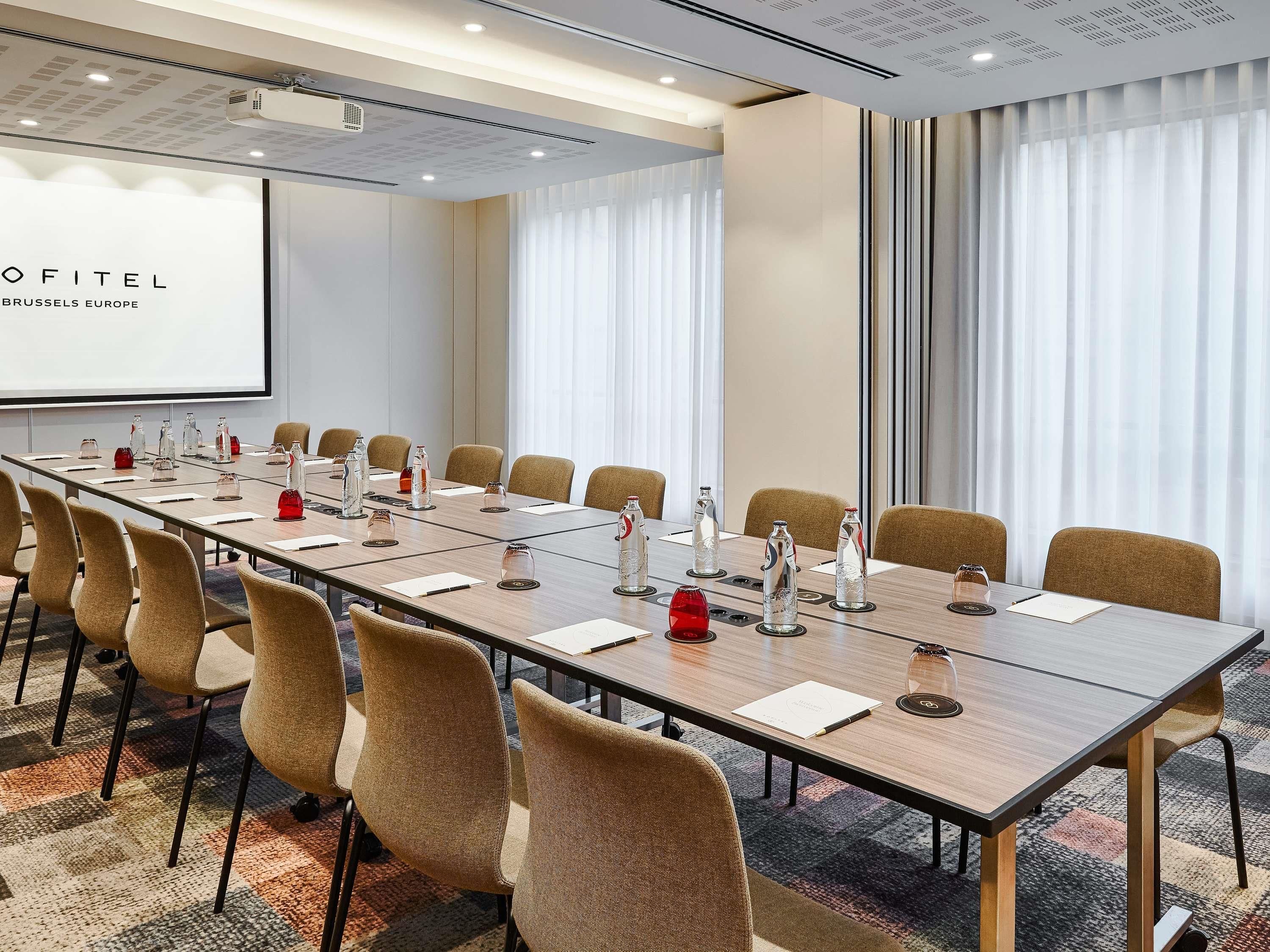 Uma sala de conferências com uma mesa comprida, cadeiras, uma tela de projetor e uma grande janela com cortinas.